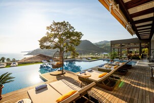 Piscina infinita com espreguiçadeiras e cadeira suspensa, com vista para uma paisagem montanhosa e o mar.
