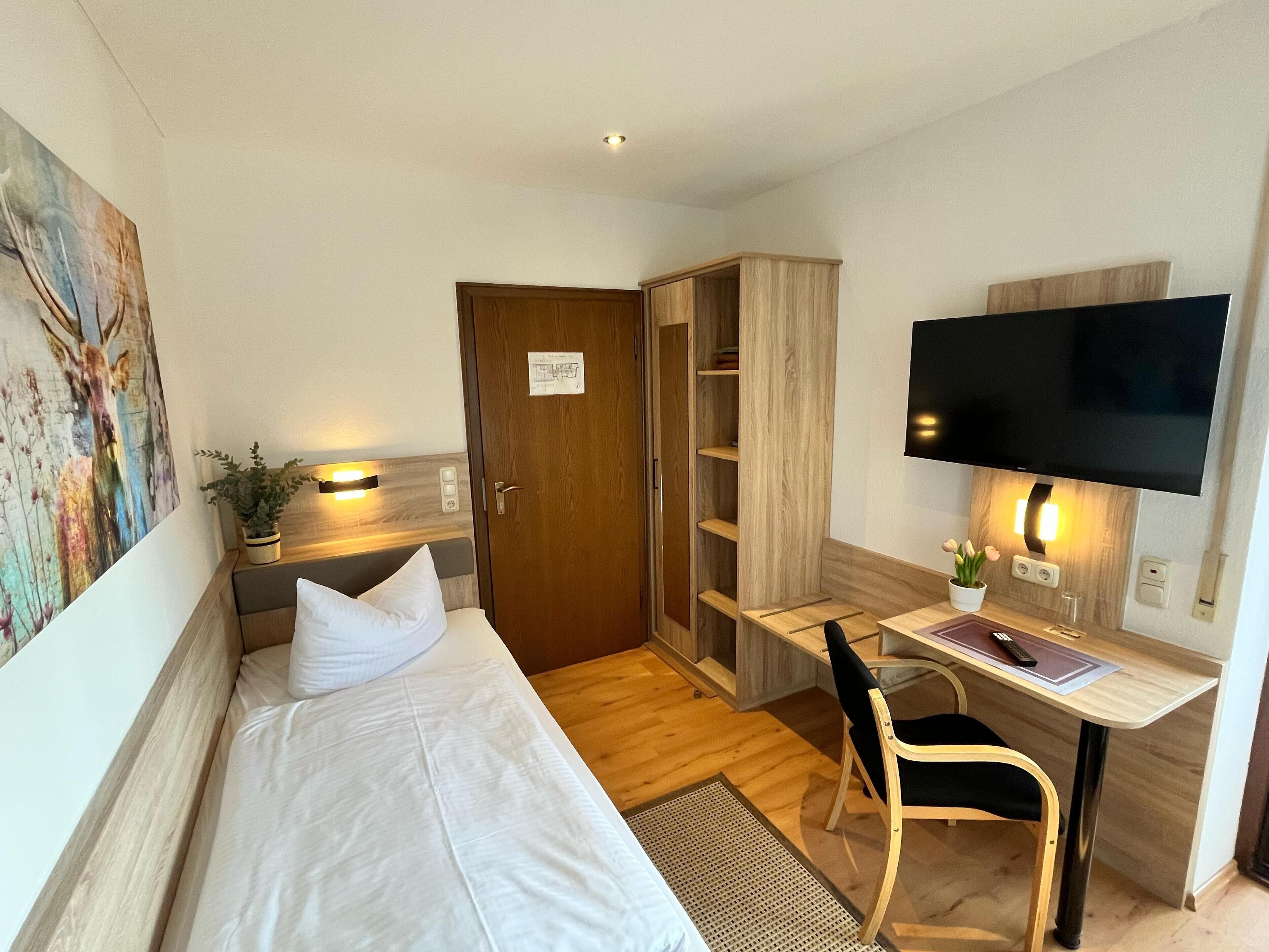 Quarto de hotel com cama, escrivaninha, cadeira, televisão, guarda-roupa e um quadro na parede.