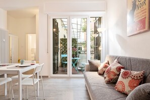 Uma sala de estar moderna com sofá, uma mesa de jantar com cadeiras e uma porta de vidro deslizante que dá para uma área externa com mesa e cadeiras.