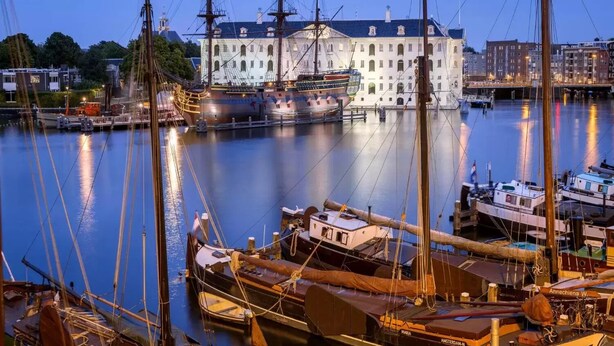 Uma marina com vários barcos à vela atracados, um edifício branco grande com janelas em arco ao fundo, e uma superfície de água calma refletindo as luzes.