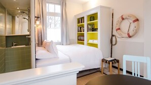 Quarto de hotel com cama, banheiro com chuveiro, estante e relógio de parede.