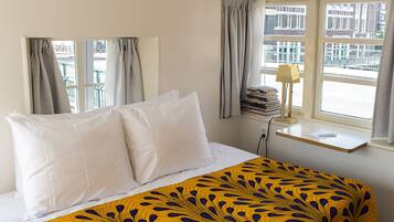 Quarto de hotel com cama, janela com cortinas, mesinha com abajur e vista para a rua.