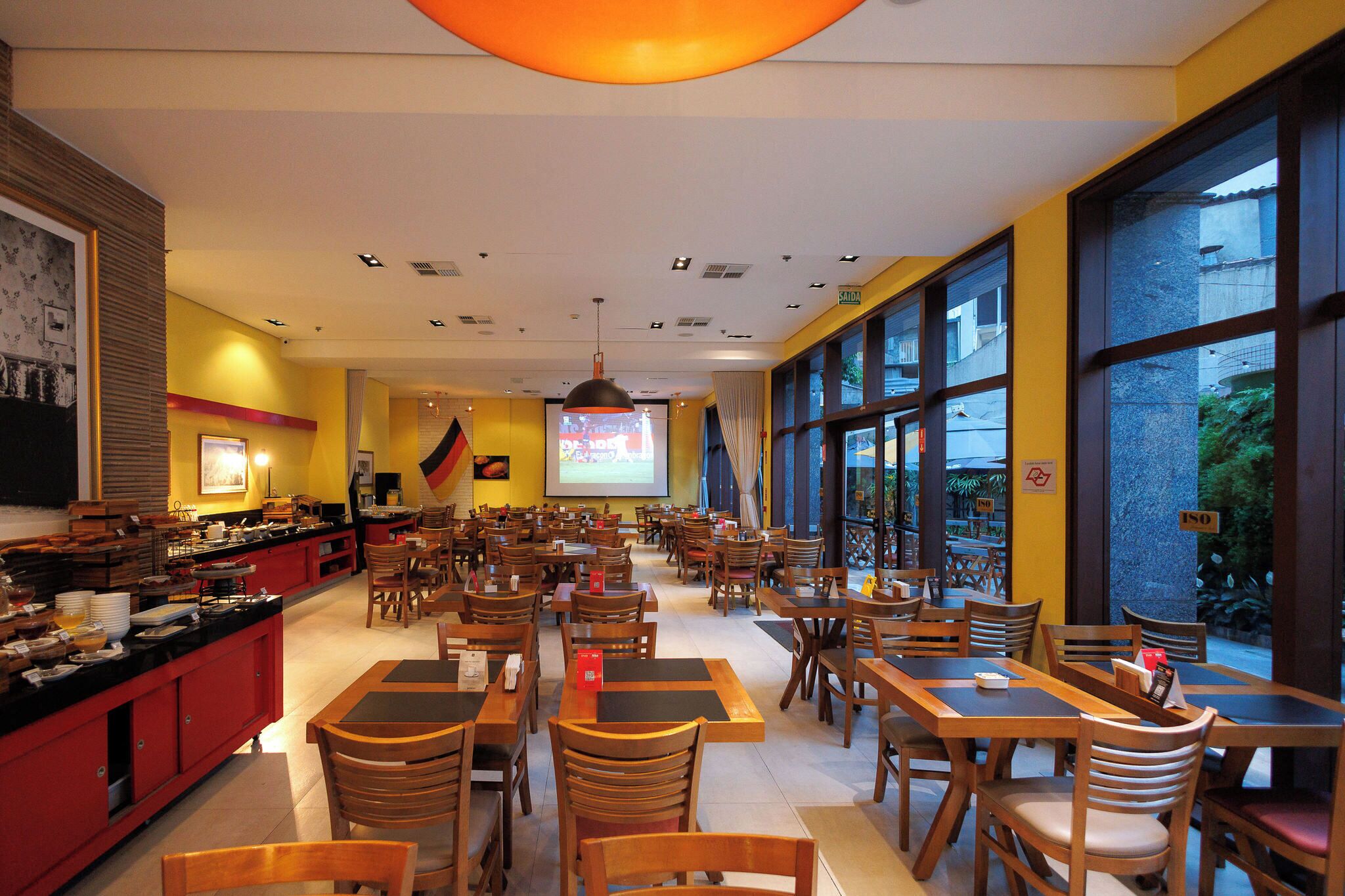 Restaurante com várias mesas, cadeiras, espaço para buffet e grandes janelas.