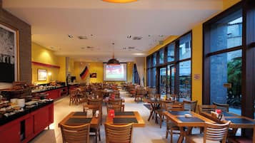 Restaurante com várias mesas, cadeiras, espaço para buffet e grandes janelas.