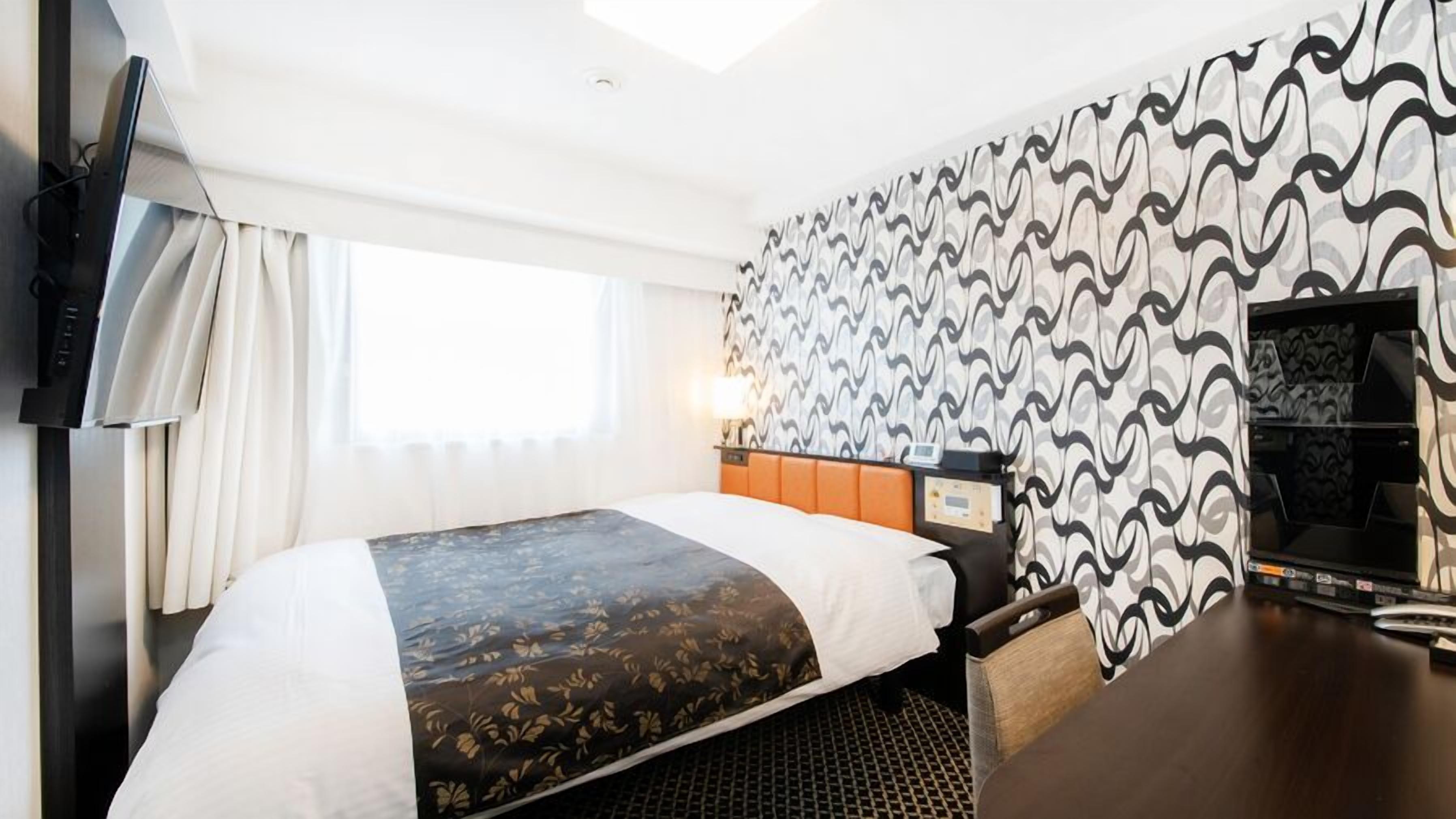 Quarto de hotel com cama, escrivaninha, televisão e parede estampada.