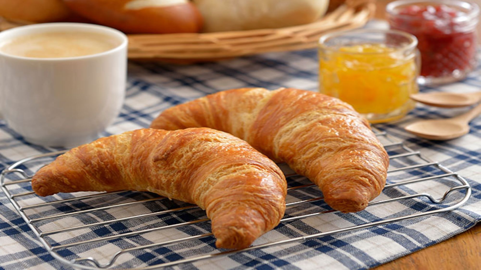 Dois croissants sobre uma grade, uma chávena de café e uma cesta com pão e geleia.