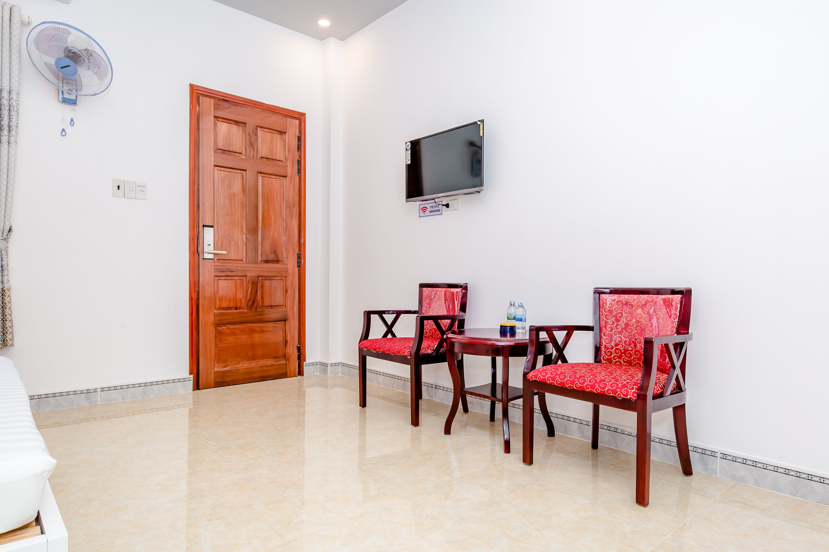 Quarto de hotel com porta de madeira, TV de tela plana na parede, duas cadeiras de madeira com almofadas vermelhas estampadas, uma mesinha e um ventilador de chão.
