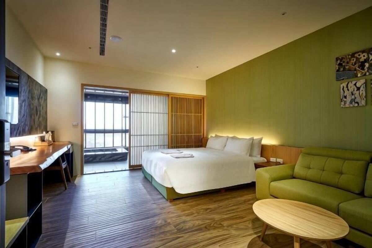 Quarto de hotel com uma cama, um sofá verde, uma escrivaninha e uma televisão.
