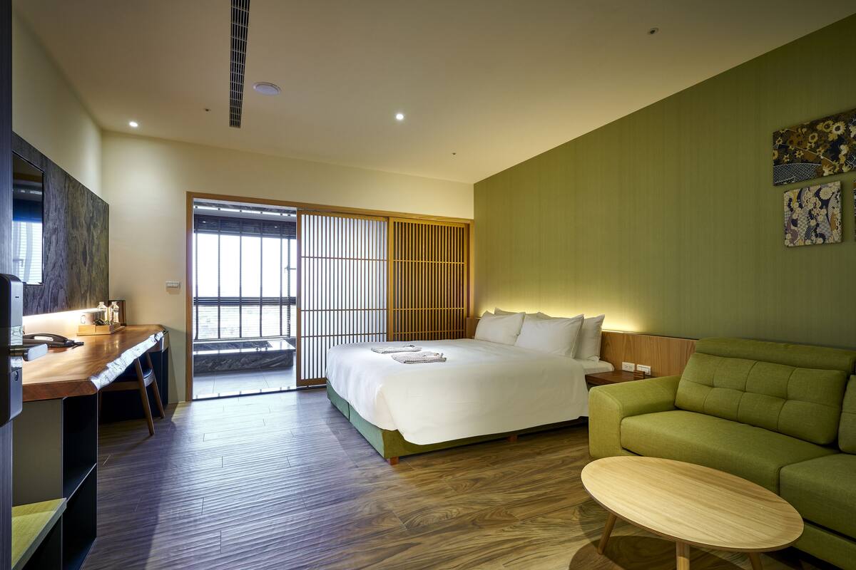 Quarto de hotel moderno com uma cama grande, um sofá verde, uma escrivaninha de madeira e uma mesa pequena.