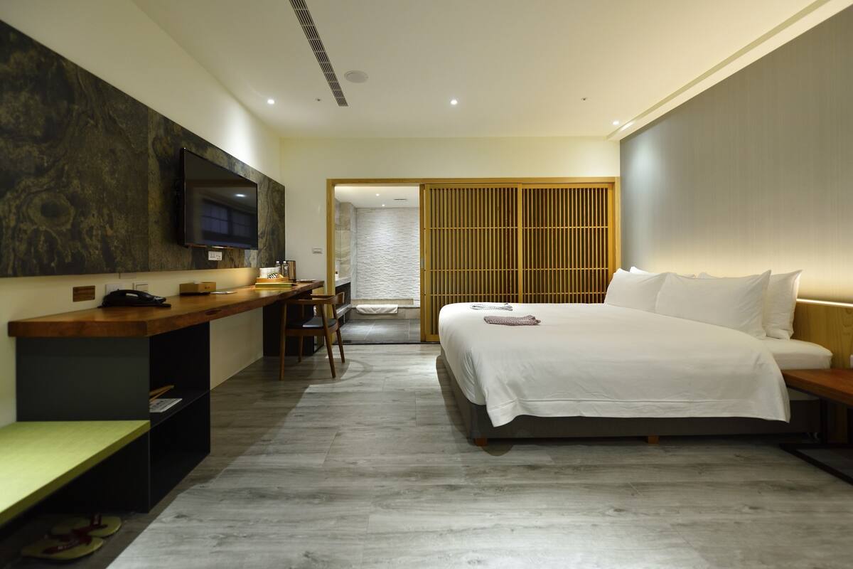 Quarto de hotel moderno com uma cama grande, uma escrivaninha com cadeira, uma TV de tela plana na parede e uma porta deslizante para um banheiro.