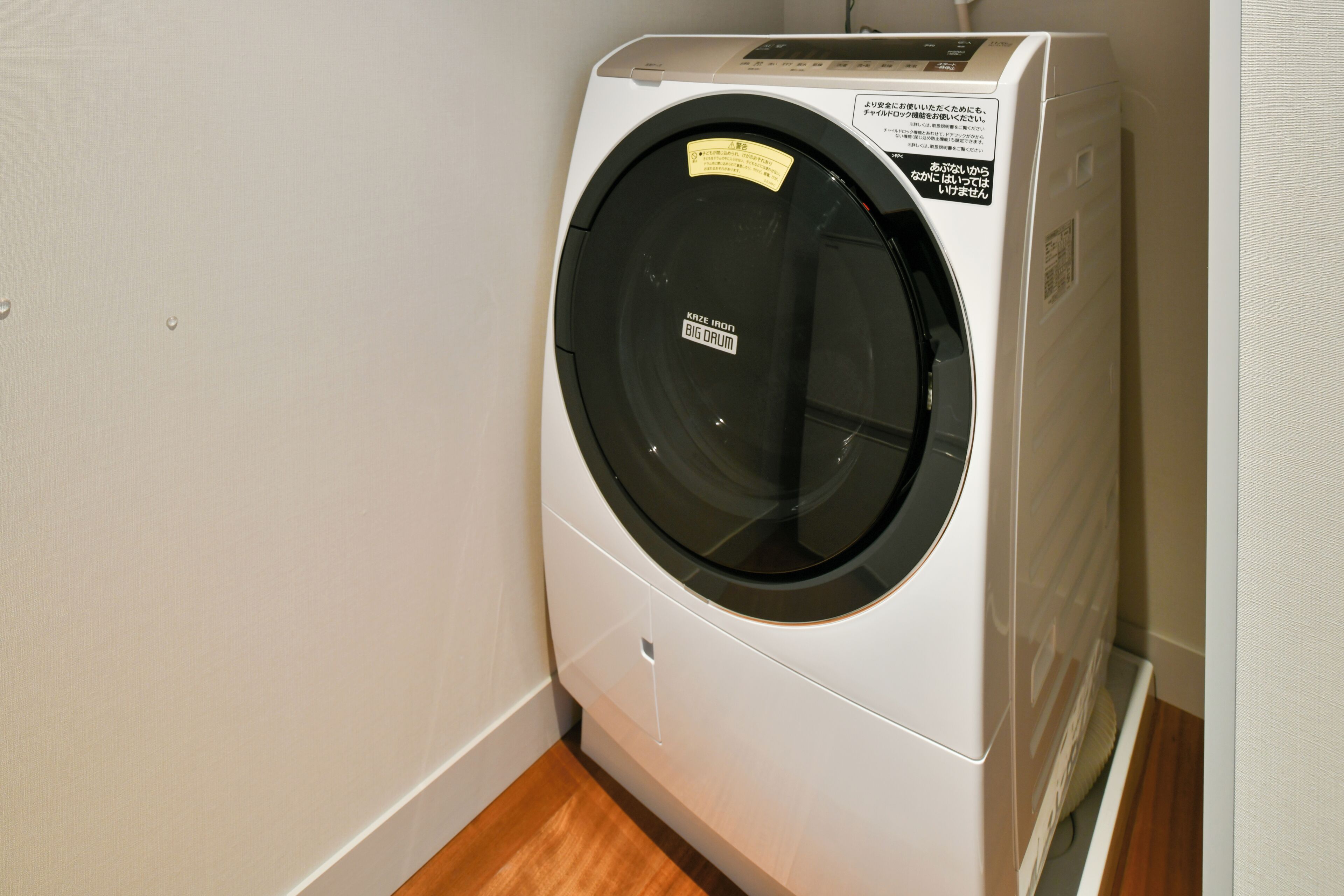 Máquina de lavar roupa compacta, de carregamento frontal e cor branca, num canto de um quarto com chão de madeira e paredes brancas.