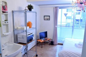 Quarto de hotel com cama, televisão, secretária com computador, prateleira com artigos de higiene pessoal, janela com cortinas e varanda.