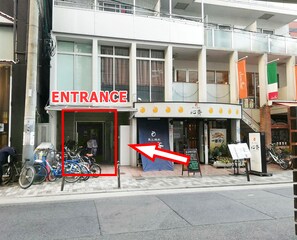 Um edifício com entrada marcada por uma placa vermelha. Bicicletas estão estacionadas do lado de fora, e uma placa com texto é visível.