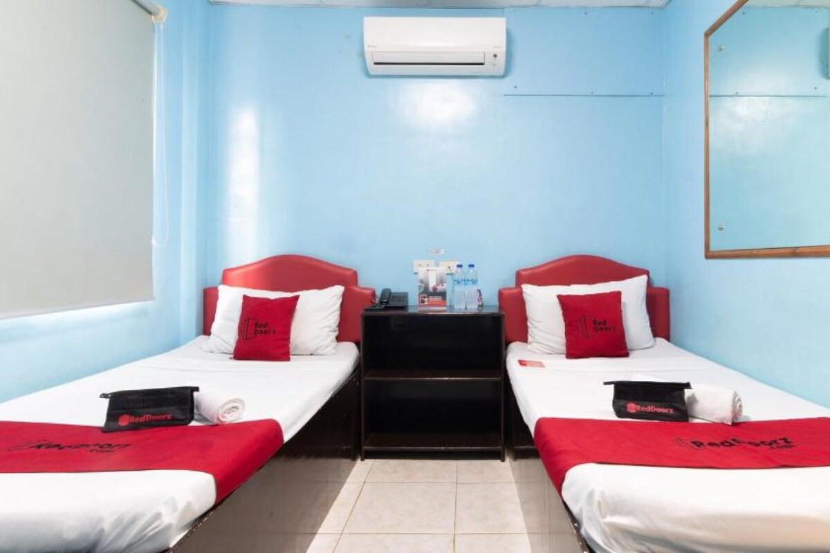 Quarto de hotel com duas camas individuais, cada uma com cabeceira vermelha e roupa de cama branca. Há uma pequena mesa com um telefone e uma garrafa de água entre as camas. O quarto tem paredes azul-claras, uma janela à esquerda e um aparelho de ar condicionado na parede.