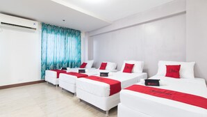 Quarto de hotel com quatro camas, cada uma com colcha vermelha e branca e almofada com o logotipo Red Doorz. Há uma janela grande com cortinas azuis, um aparelho de ar condicionado na parede e piso de madeira.