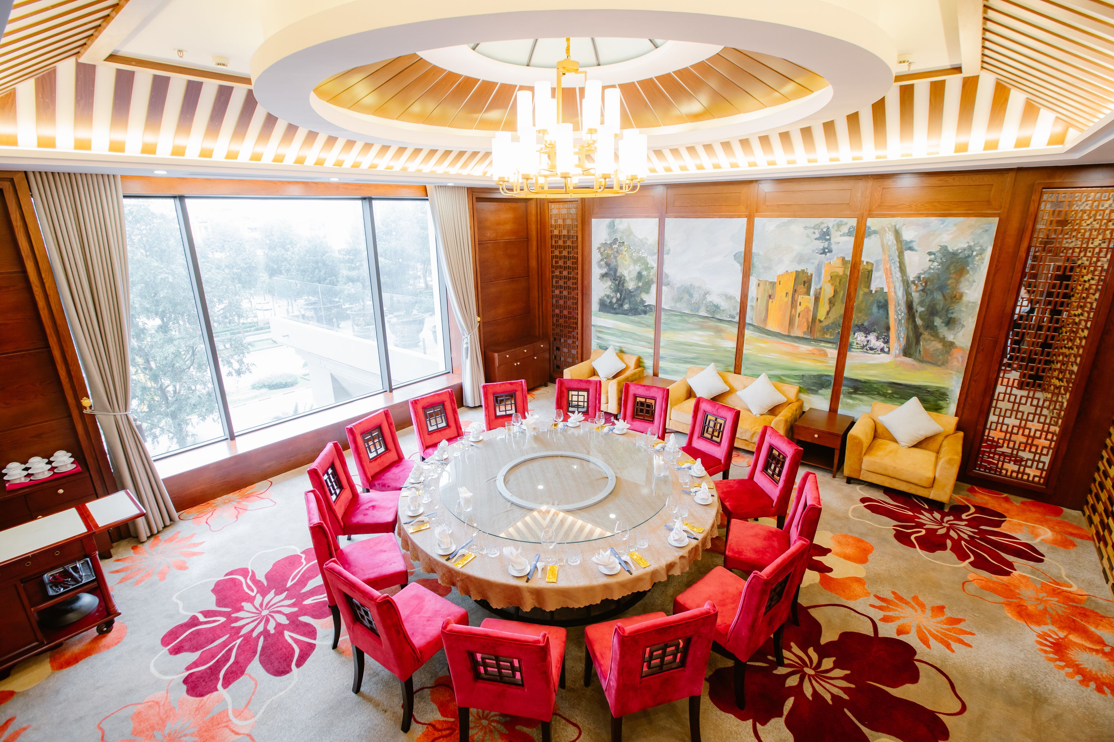 Uma mesa redonda com cadeiras vermelhas, um candelabro e uma grande janela com vista para árvores e edifícios.