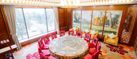 Uma mesa redonda com cadeiras vermelhas, um candelabro e uma grande janela com vista para árvores e edifícios.