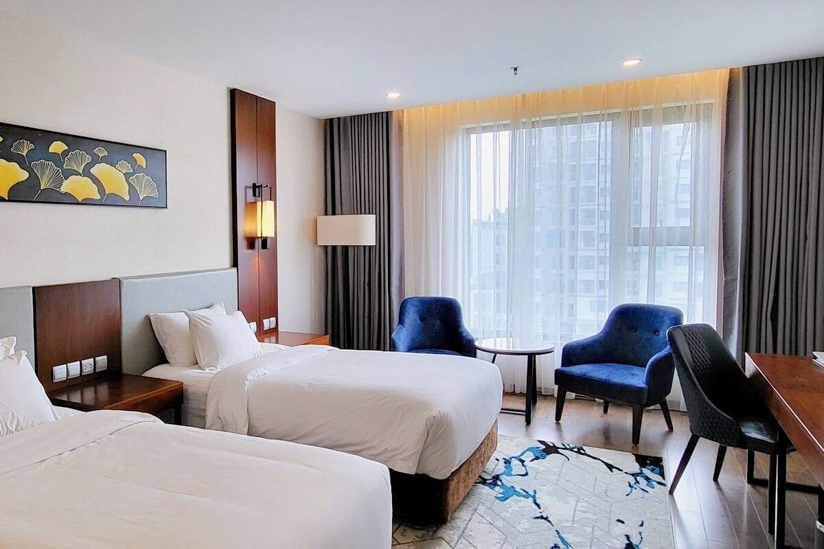 Quarto de hotel com duas camas, uma escrivaninha, uma cadeira, uma luminária, um quadro e uma janela grande com cortinas.