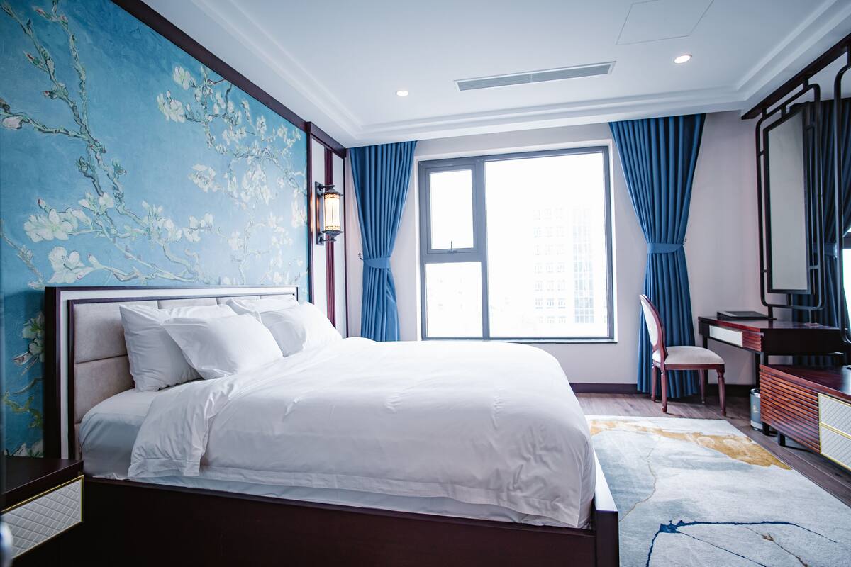 Quarto de hotel com uma cama grande, uma mesa, uma cadeira e uma janela com cortinas.