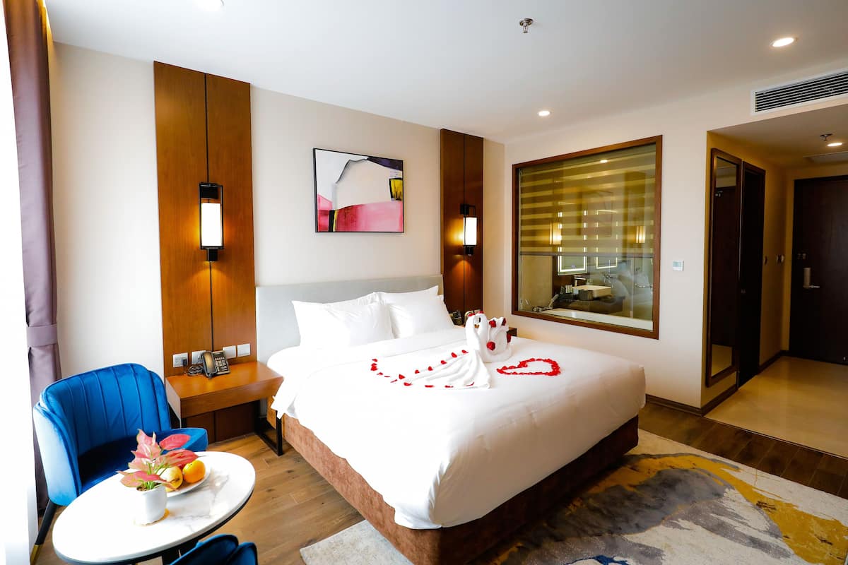Quarto de hotel com uma cama grande, uma cadeira azul, uma mesinha com uma planta e um quadro na parede.