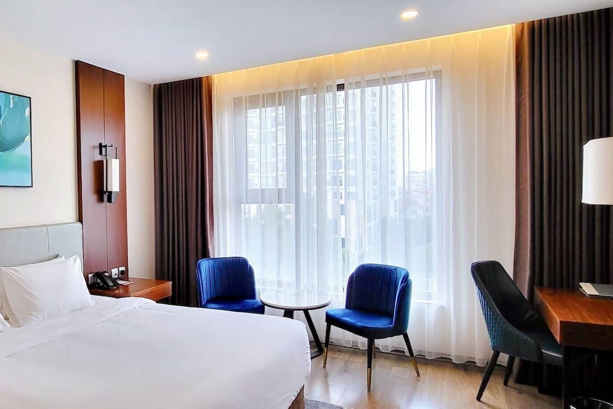 Quarto de hotel com uma cama grande, duas cadeiras azuis, uma escrivaninha, uma televisão e um quadro na parede.