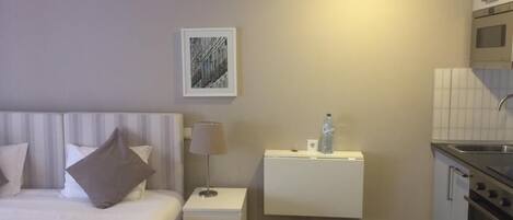 Quarto de hotel com cama, mesa de cabeceira, abajur, garrafa de água e cozinha equipada com fogão e forno.