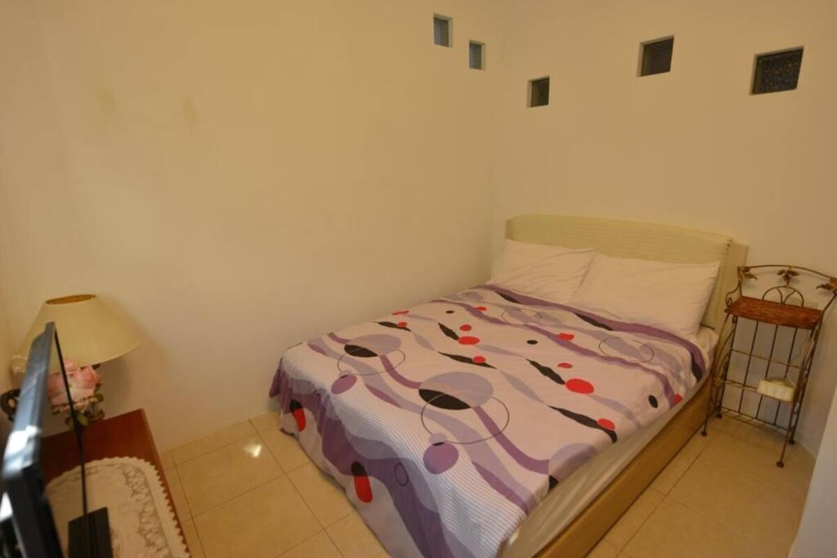 Quarto com cama, mesa de cabeceira, abajur e uma pequena mesa com uma flor.
