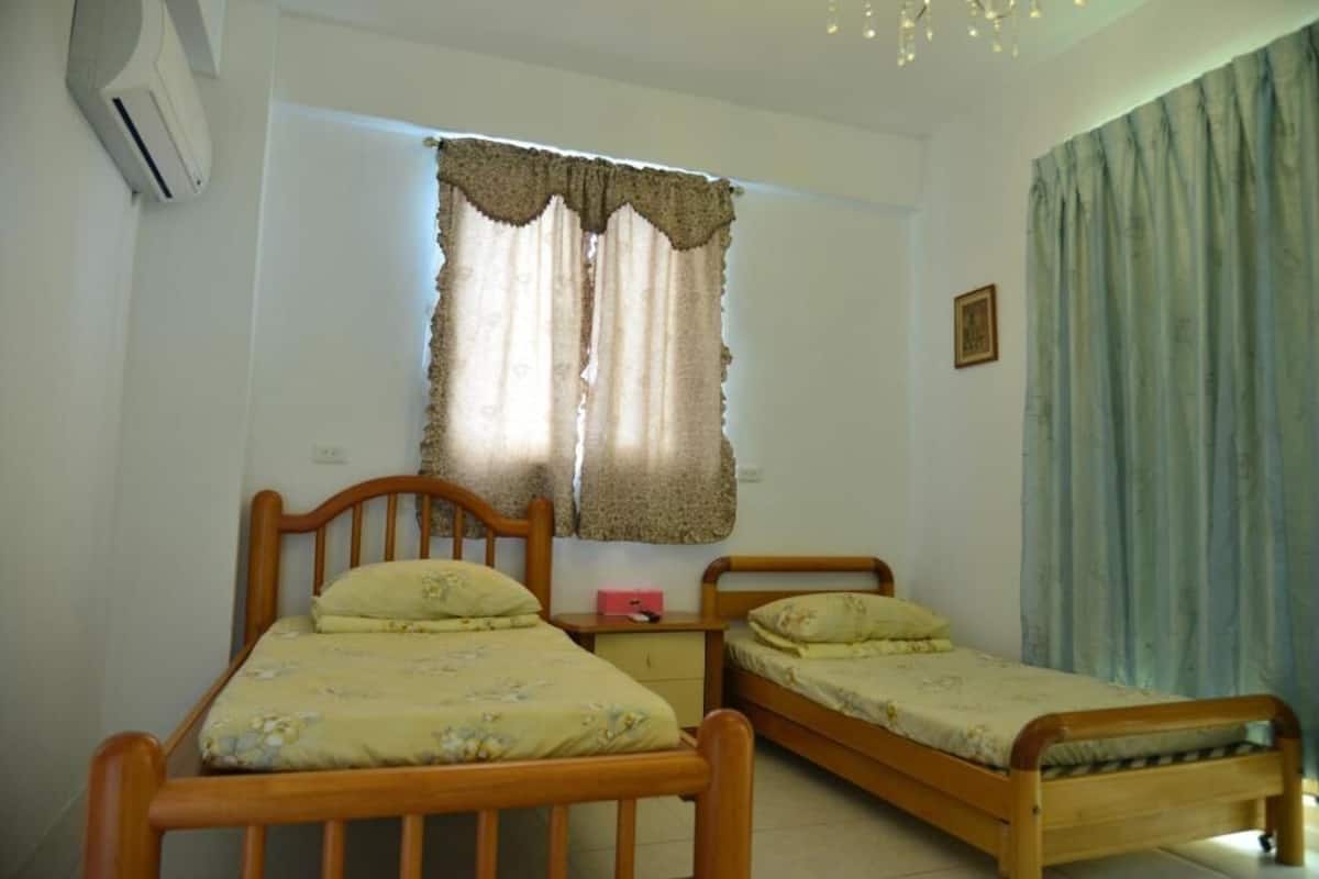 Quarto com duas camas de madeira, janela com cortinas, mesa de cabeceira e ar condicionado fixado na parede.