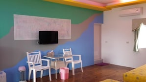 Quarto com parede azul e branca, televisão de tela plana sobre uma pequena mesa com duas cadeiras brancas, cama amarela e janela com cortinas.