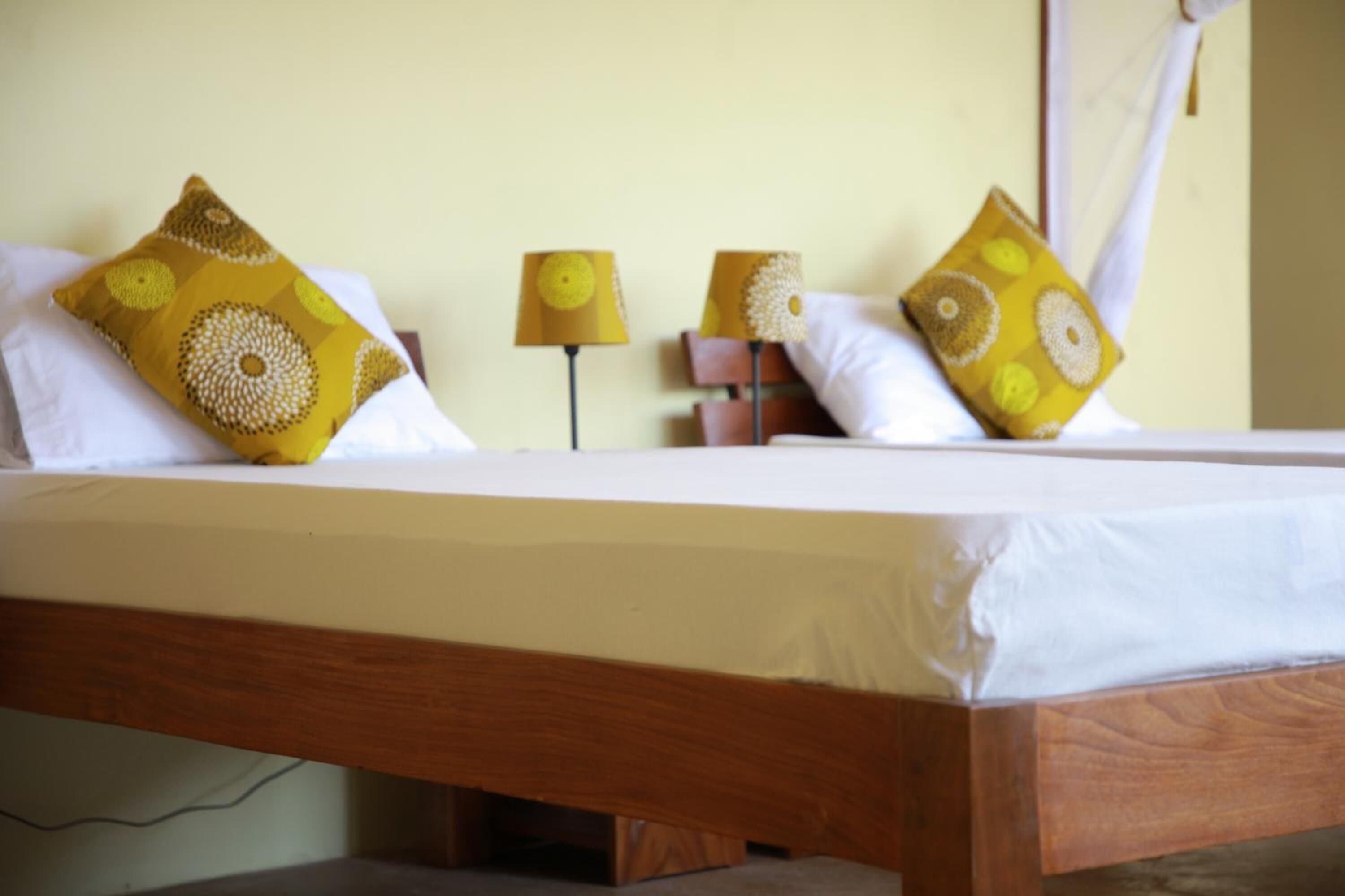 Uma cama com estrutura de madeira, duas almofadas com padrão amarelo e branco, e duas luminárias de cabeceira com abajures amarelos.
