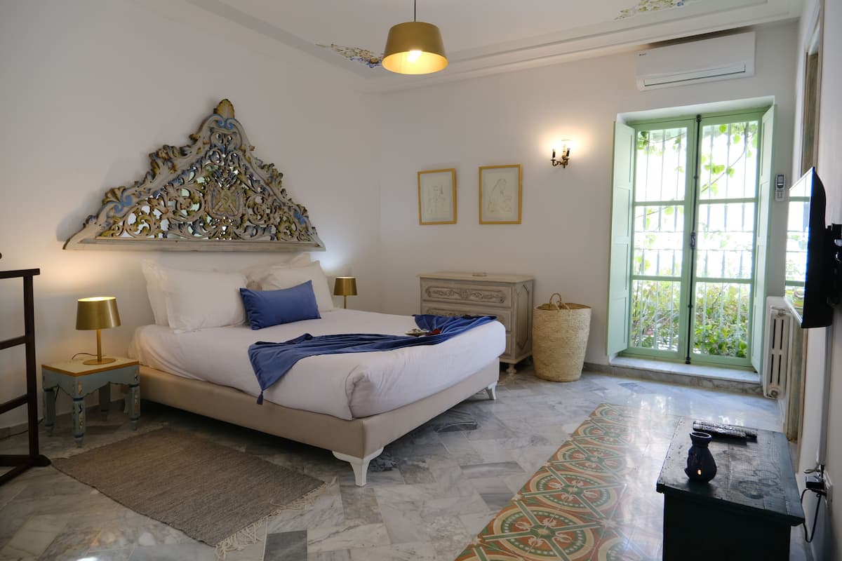 Quarto com cama, mesas de cabeceira, um painel de cabeceira decorativo, janela com vista e piso estampado.