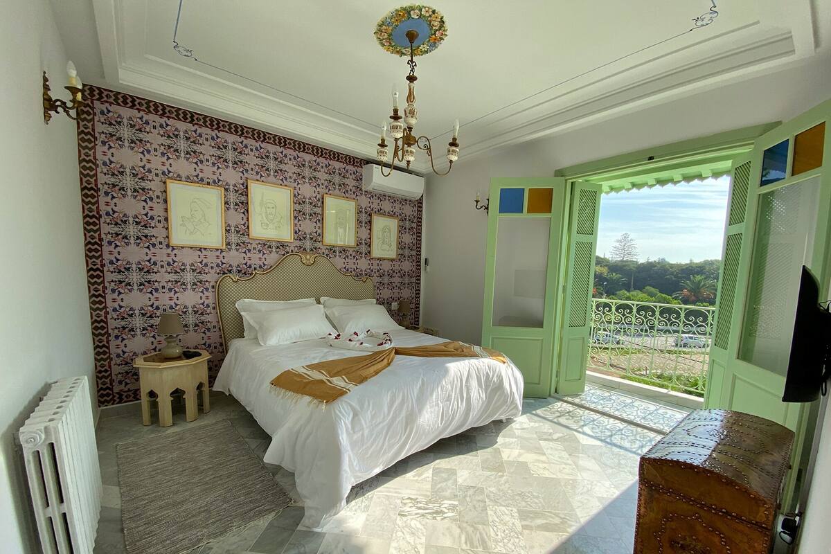 Quarto com cama, radiador, mesinha de cabeceira com abajur, cômoda e varanda com vista.