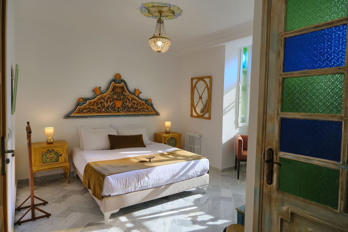 Quarto com cama grande, duas mesas de cabeceira com luminárias, cabeceira decorativa, radiador e janela com vidros verdes.