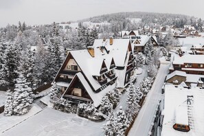 Uma aldeia coberta de neve com casas de madeira tradicionais, um edifício central com varanda e um cenário de floresta.