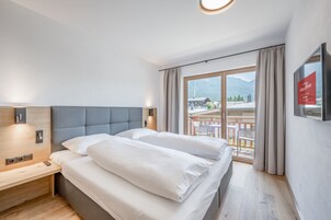 Quarto de hotel moderno com uma cama grande, mesa de cabeceira de madeira, televisão de tela plana e varanda com vista para as montanhas.