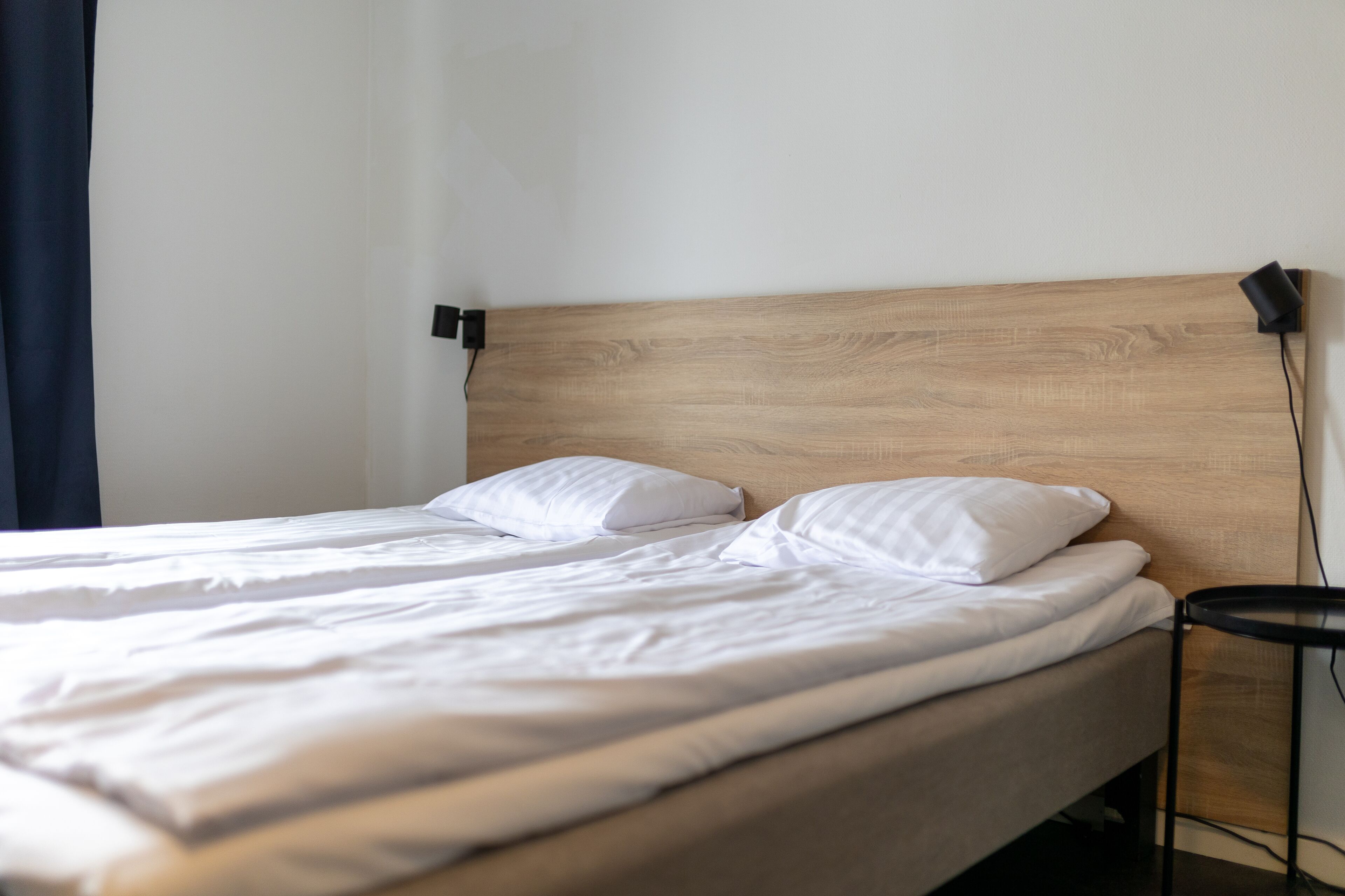 Cama bem arrumada com lençóis brancos e duas almofadas, cabeceira de madeira e uma pequena mesa de cabeceira preta.
