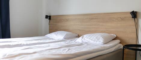 Cama bem arrumada com lençóis brancos e duas almofadas, cabeceira de madeira e uma pequena mesa de cabeceira preta.