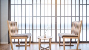 Duas cadeiras de madeira com almofadas, uma mesa redonda pequena com um vaso e uma janela grande com persianas verticais.
