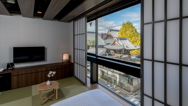 Quarto de hotel moderno com uma janela grande, televisão de tela plana, mesa de cabeceira de madeira e uma porta japonesa tradicional de correr.