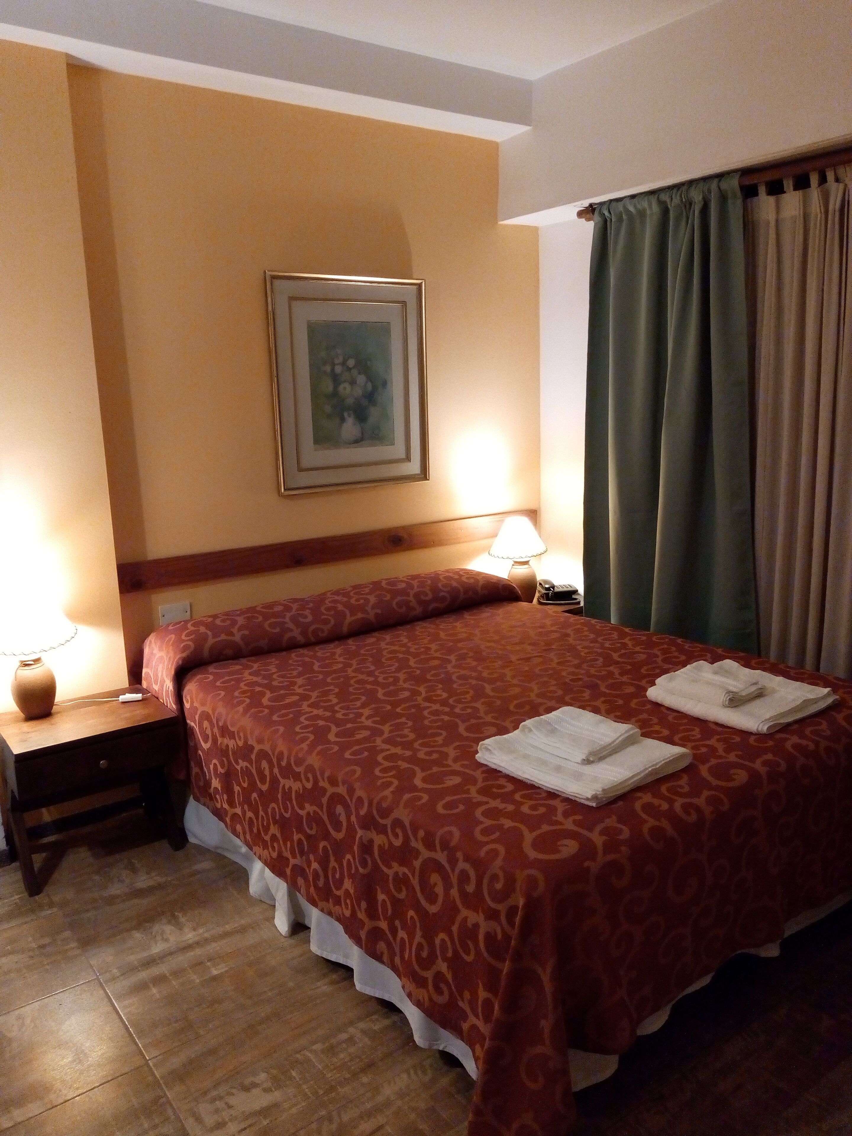 Quarto de hotel com uma cama grande, duas luminárias de cabeceira, um quadro na parede e uma janela com cortinas.