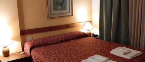 Quarto de hotel com uma cama grande, duas luminárias de cabeceira, um quadro na parede e uma janela com cortinas.