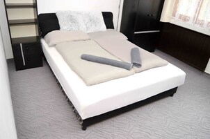 Cama bem arrumada com colchão branco, manta bege e toalha cinza enrolada. Um criado-mudo preto com gaveta e uma luminária pequena ficam ao lado da cama. O quarto tem um tapete com padrão herringbone e um guarda-roupa de madeira escura ao fundo.