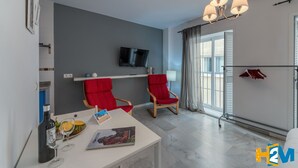Uma sala de estar moderna com uma TV de tela plana, duas cadeiras vermelhas, uma mesa de jantar com garrafa e copos, e uma janela grande com cortinas.