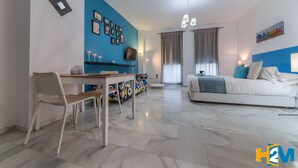 Quarto de hotel moderno com parede azul de destaque, sofá branco, mesa de jantar com cadeiras e uma TV.