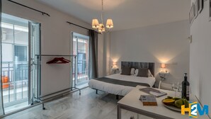 Quarto de hotel moderno com uma cama grande, abajures, uma mesa com uma garrafa de vinho e frutas, e uma varanda com um varal.