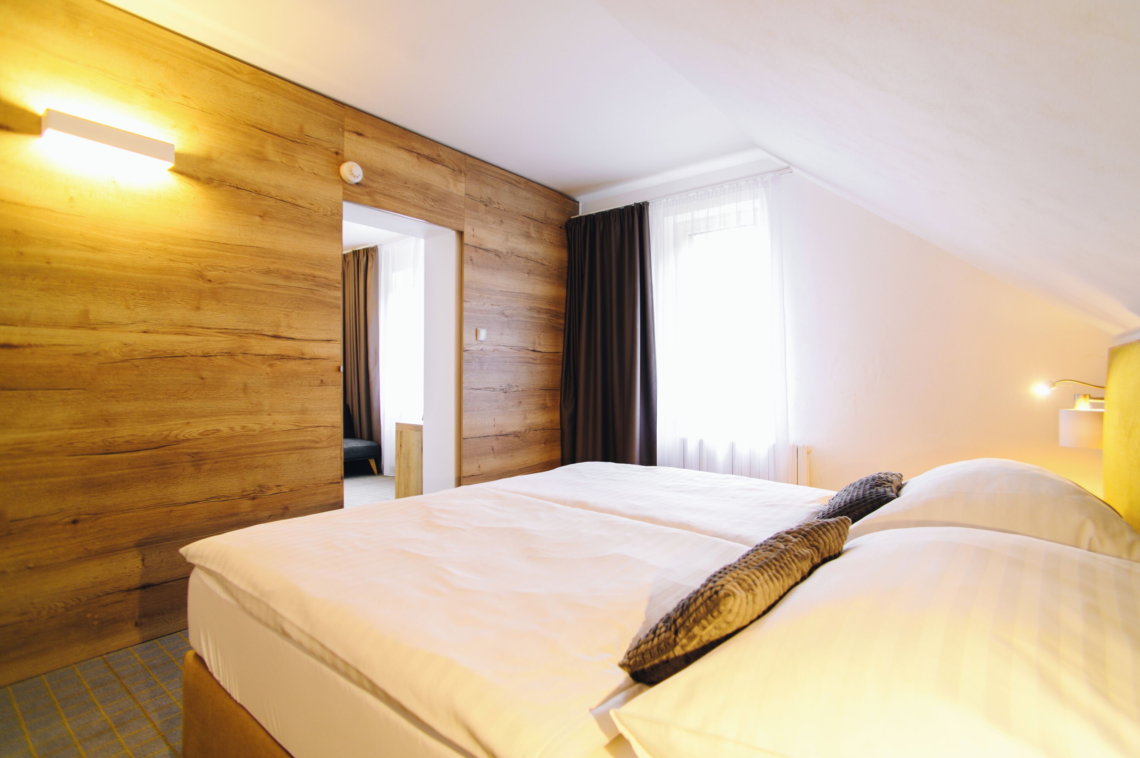 Quarto de hotel com cabeceira de madeira, uma cama grande com lençóis brancos, uma janela com cortinas e uma luminária de cabeceira.