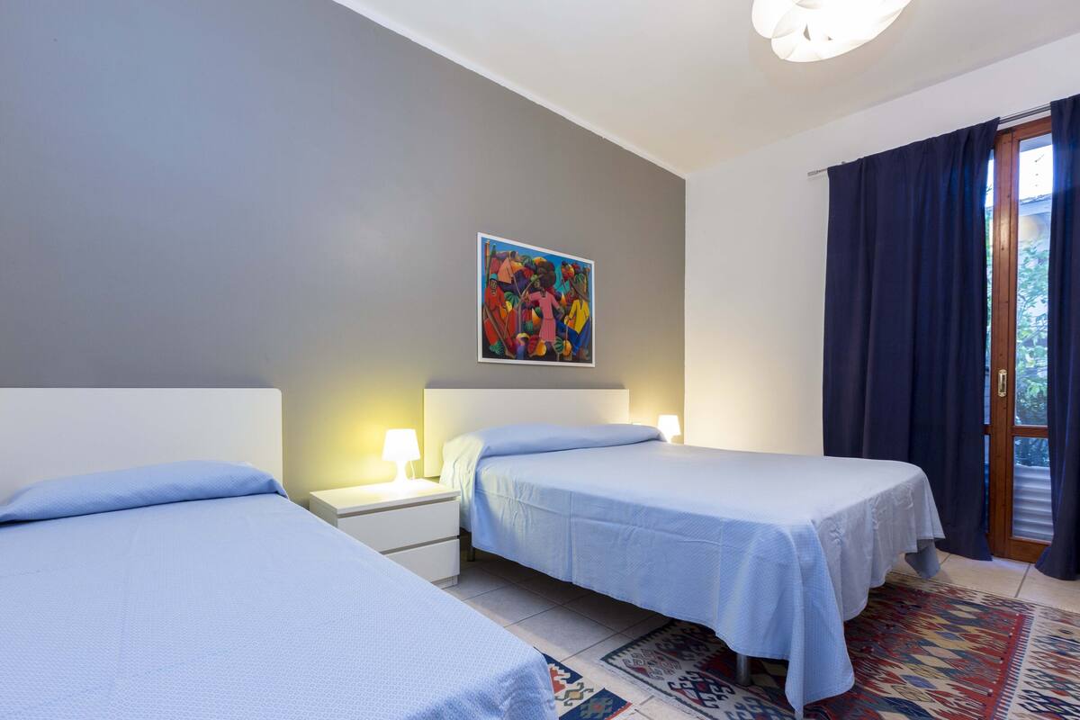 Quarto de hotel com duas camas, um quadro na parede, uma mesa de cabeceira com abajur e vista através de uma porta com cortinas.
