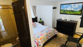 Quarto de hotel com uma cama, uma escrivaninha, uma cadeira, um quadro na parede e um banheiro com vaso sanitário e chuveiro.
