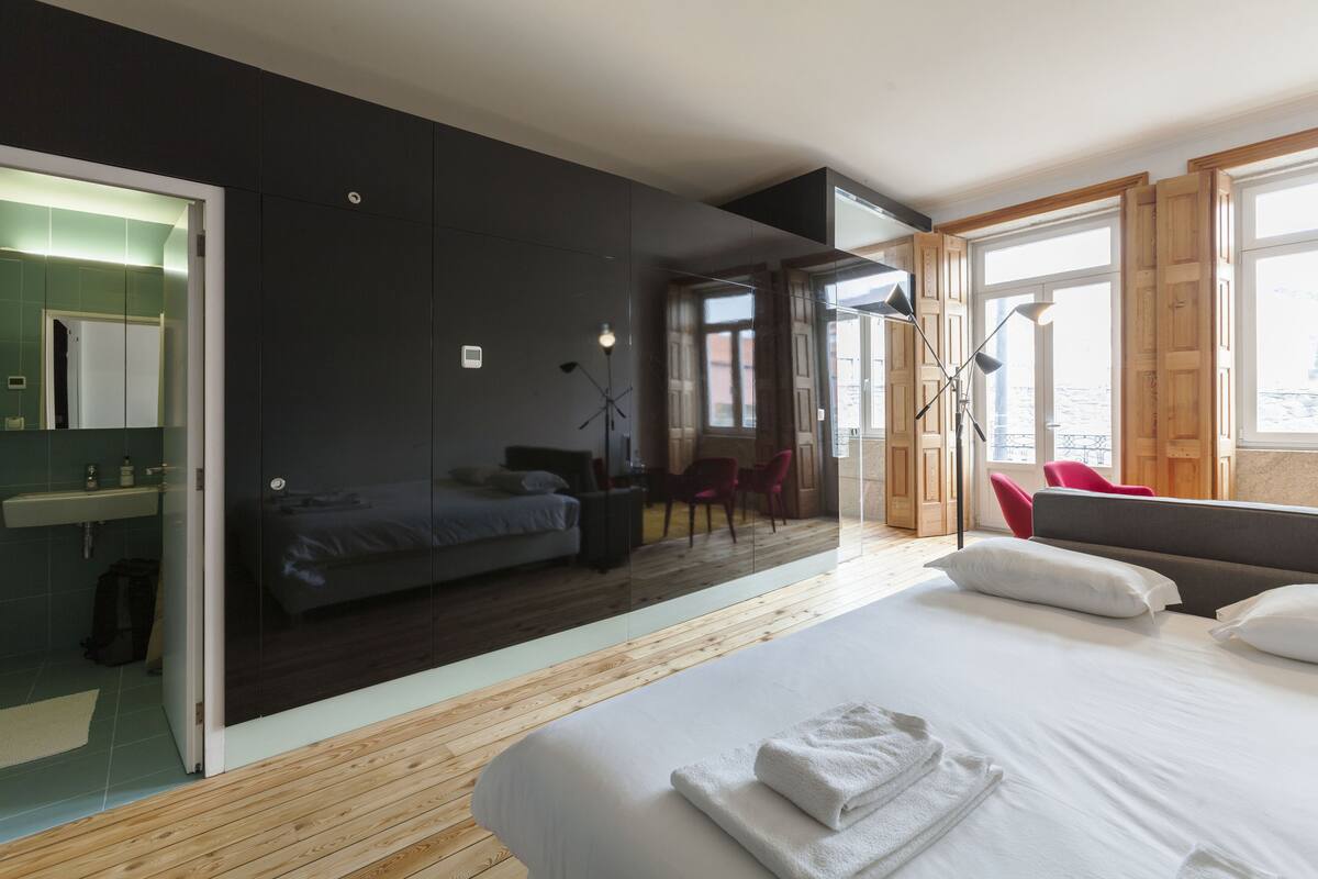 Quarto de hotel moderno com uma cama grande, área de estar separada com uma poltrona, escrivaninha e uma janela ampla com vista.