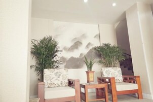Uma área de estar com duas cadeiras, uma mesa, um quadro na parede e duas plantas em vasos.